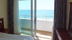 Quarto de hotel com cama, escrivaninha e vista para o mar através de uma porta de vidro deslizante.