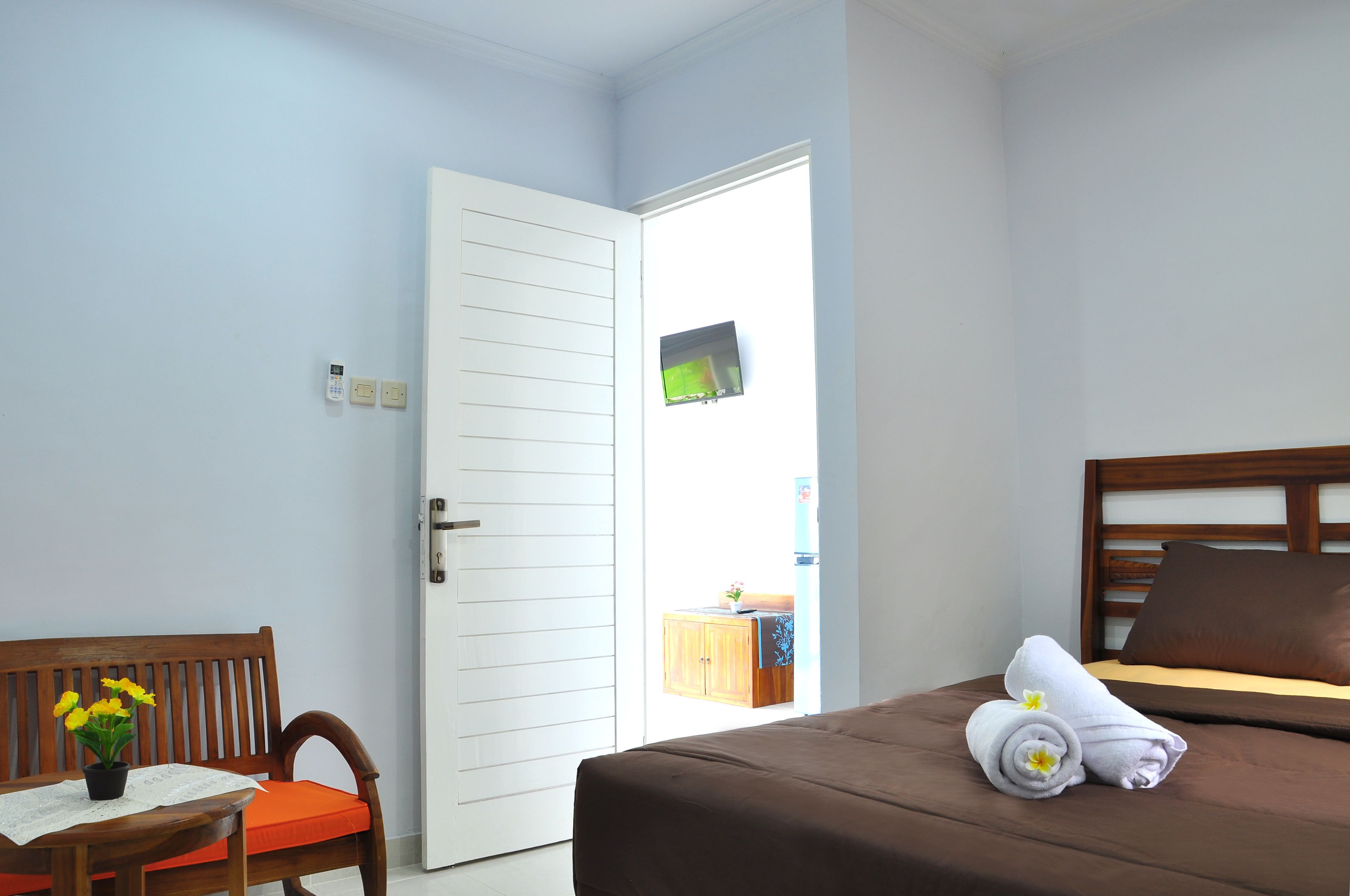 Quarto de hotel com uma cama de madeira, uma mesa pequena com planta em vaso, um banco e uma porta que dá para um banheiro.