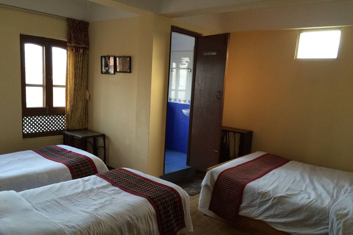Quarto de hotel com duas camas, uma pequena mesa, uma janela com cortinas, uma porta que dá para o banheiro e um pequeno armário com produtos de higiene.