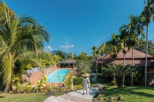 Um resort tropical com piscina, palmeiras e uma pessoa caminhando em direção à piscina.