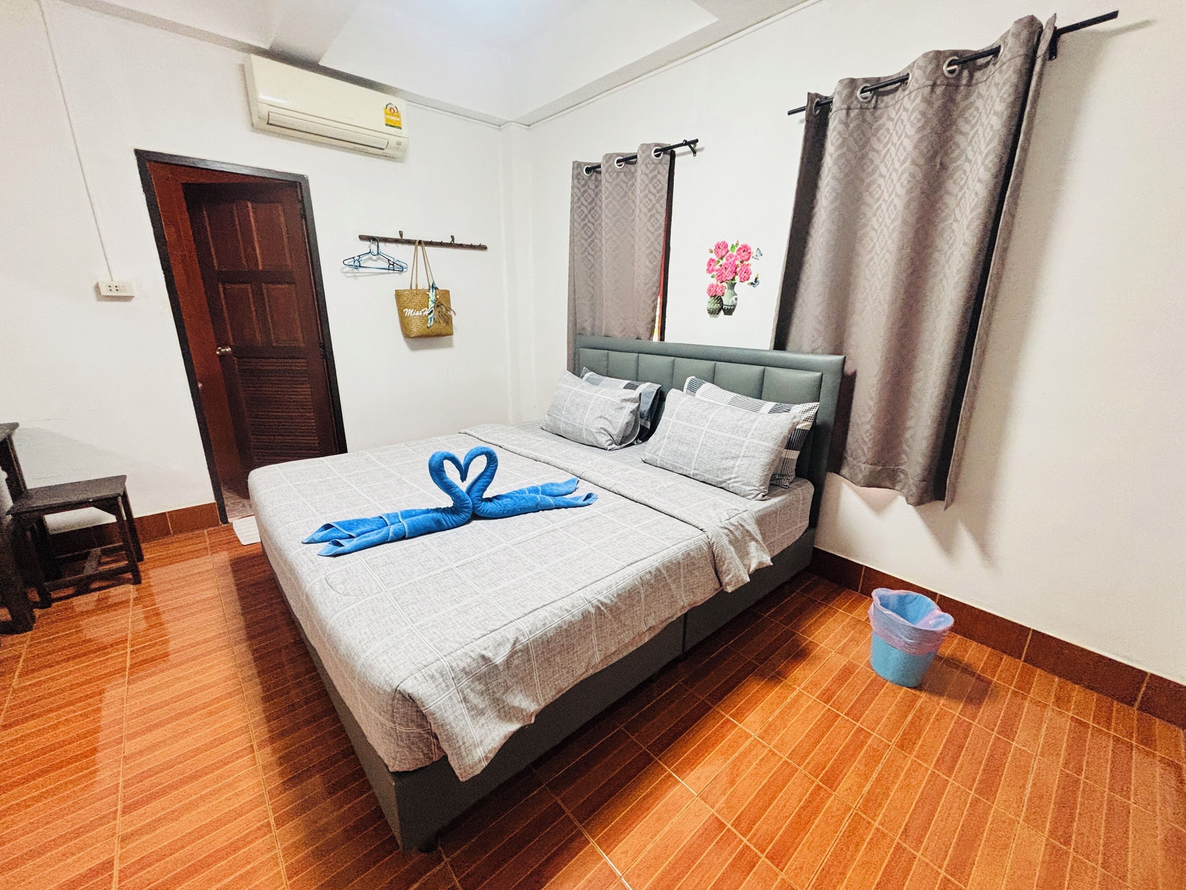 Uma cama bem arrumada com uma toalha em formato de coração, uma cadeira de madeira, uma mesa pequena, um cesto de lixo e um ar-condicionado fixado na parede.