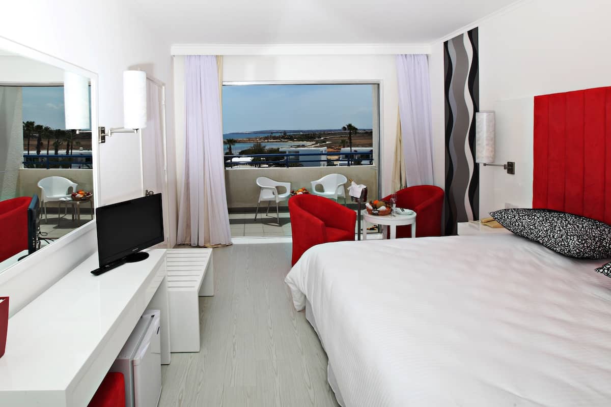 Quarto de hotel com cama, escrivaninha com computador, televisão, cadeira, mesa pequena com cesta de frutas e vista para o mar e edifícios.