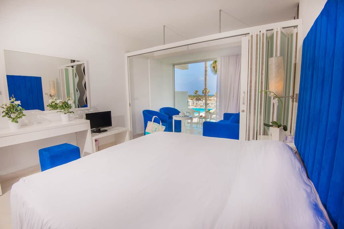 Quarto de hotel moderno com cama, escrivaninha, poltrona, televisão e varanda com vista para a piscina.