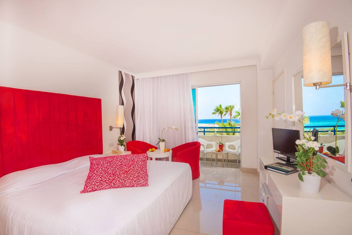 Quarto de hotel com cama, escrivaninha com televisão e varanda com vista para o mar.