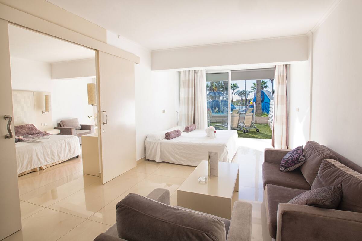Quarto de hotel moderno com cama, sofá, mesa de centro e varanda com vista para a piscina.