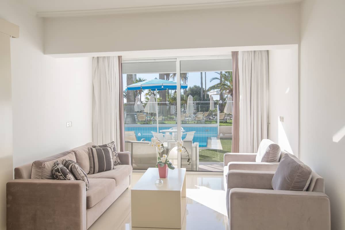 Uma sala de estar moderna com um sofá bege, uma mesa de centro e vista para uma piscina e móveis de exterior através de grandes janelas.