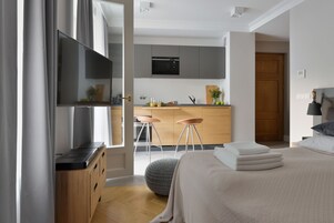Quarto moderno com uma cama grande, uma TV na parede e uma kitchenette ao fundo.