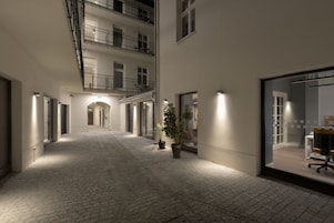 Um pátio estreito e de calçada portuguesa, com um arco central, ladeado por edifícios com varandas e grandes janelas.