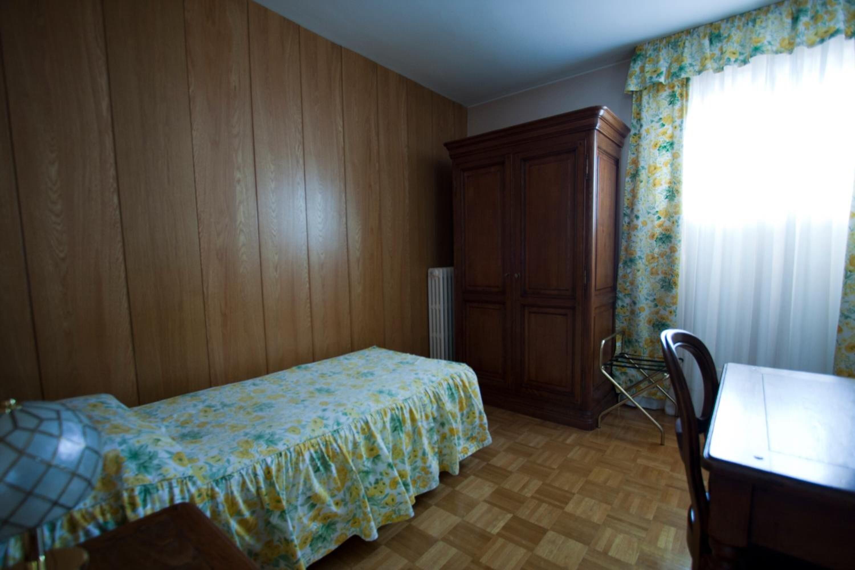 Um quarto com paredes revestidas de madeira, uma cama com colcha florida, um armário de madeira, um radiador e uma janela com cortinas florais.