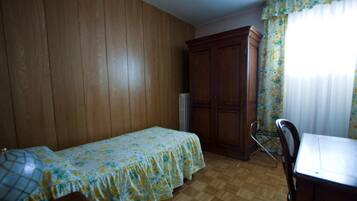 Um quarto com paredes revestidas de madeira, uma cama com colcha florida, um armário de madeira, um radiador e uma janela com cortinas florais.