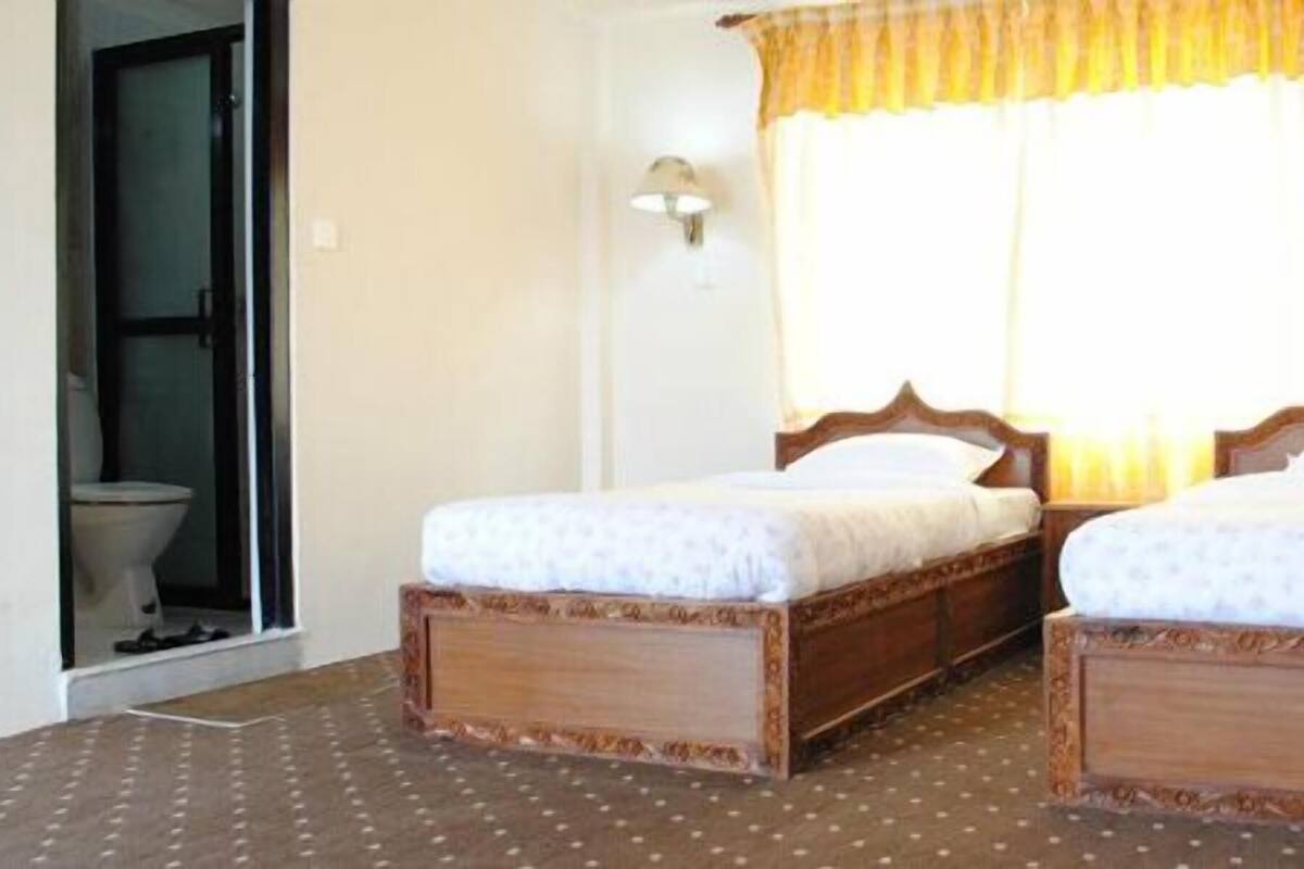 Quarto com duas camas de madeira, um toalete visível por uma porta aberta e um chão estampado.