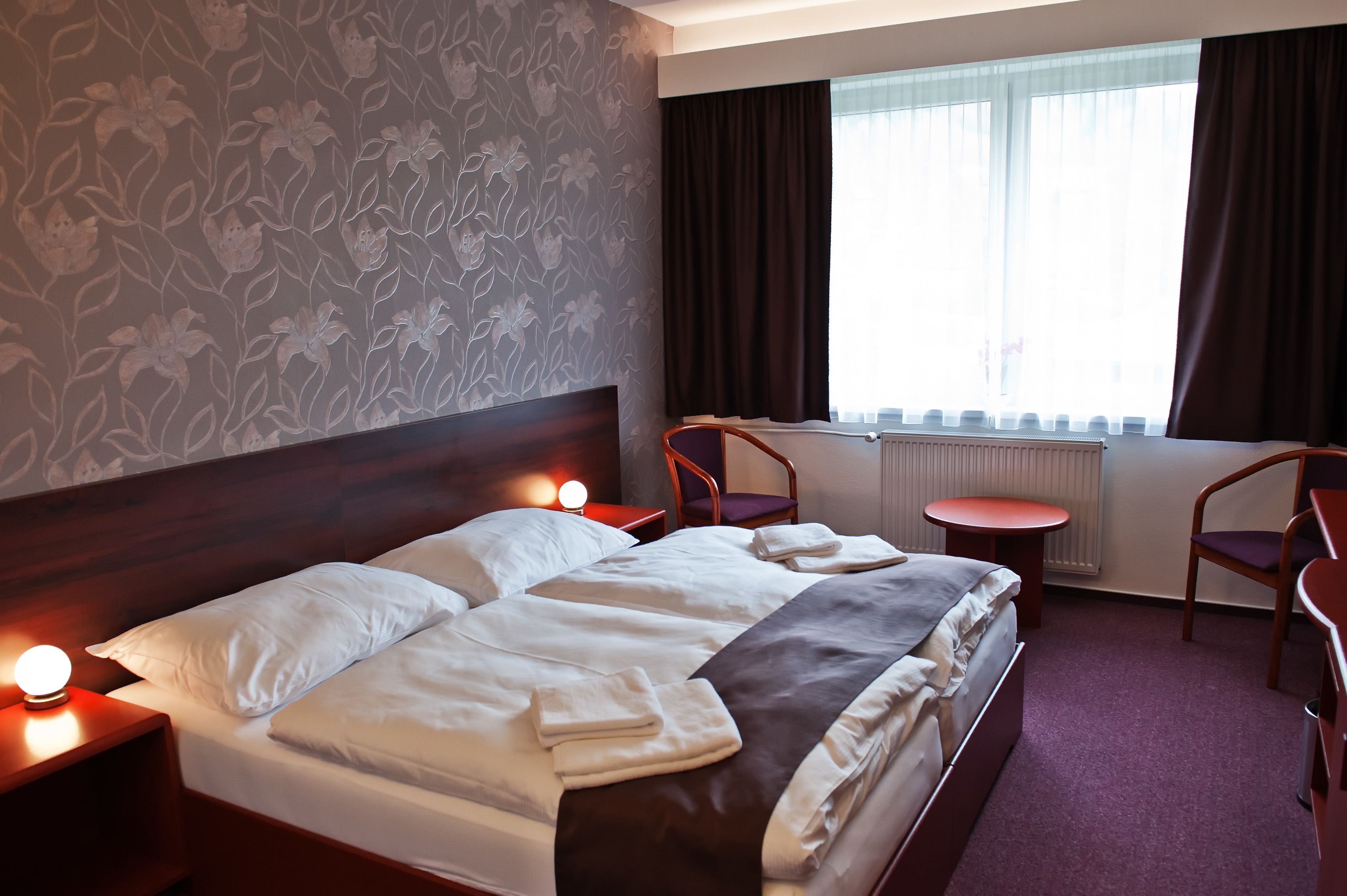 Quarto de hotel com uma cama grande, duas mesas de cabeceira com luminárias, uma mesa redonda pequena, duas cadeiras e uma janela com cortinas.
