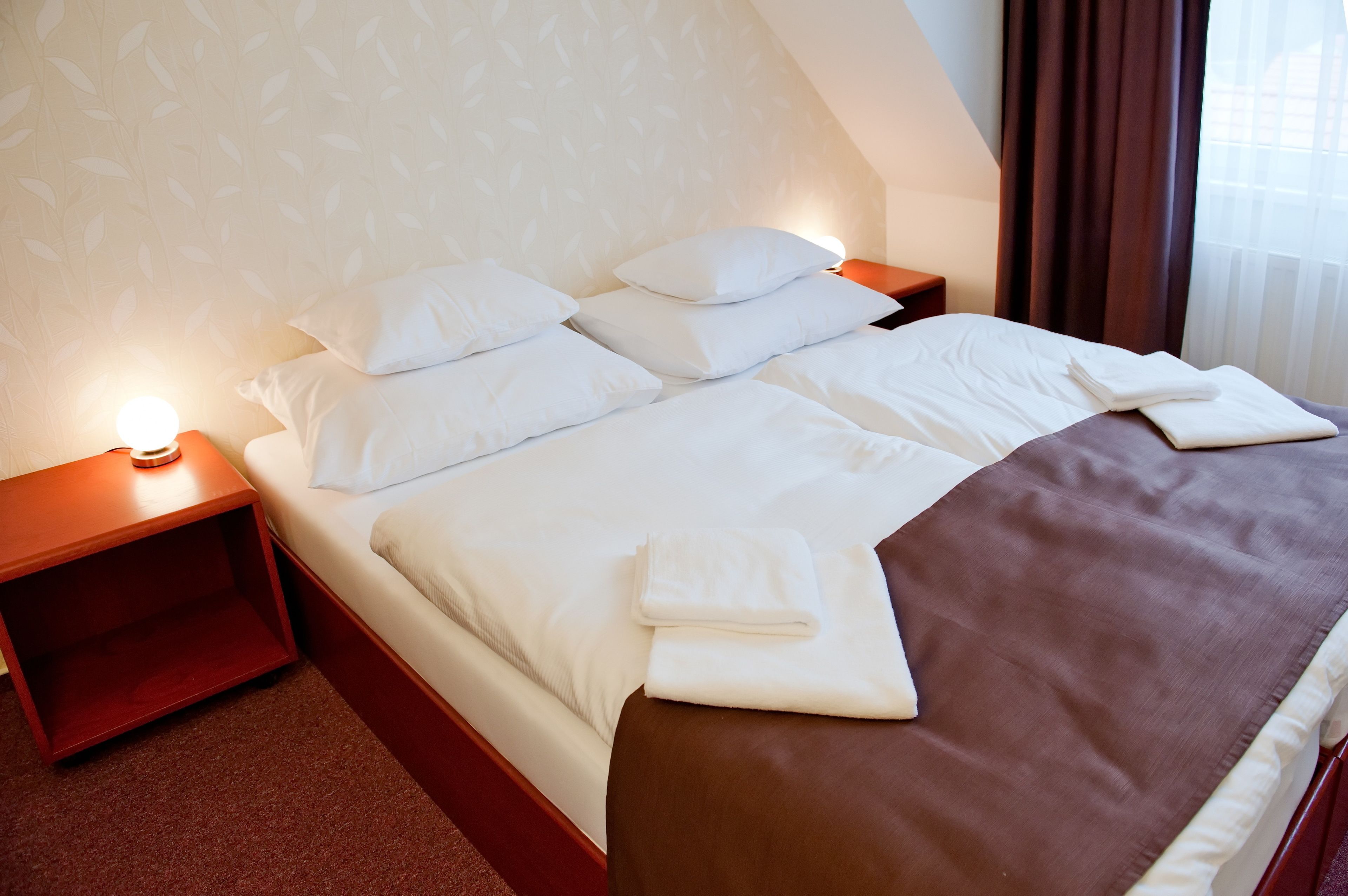 Uma cama bem arrumada com lençóis brancos e um manta marrom, um criado-mudo de madeira com uma luminária, e uma parede com padrão.