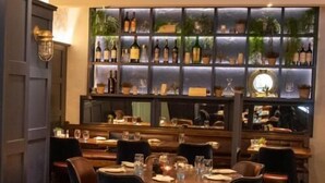 Interior de restaurante com mesas e cadeiras de madeira, área de bar com prateleiras de garrafas e uma parede decorativa com plantas.