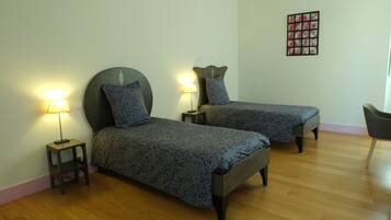 Quarto com duas camas de solteiro, cada uma com mesa de cabeceira e luminária. O quarto possui piso de madeira e uma obra de arte pendurada na parede.