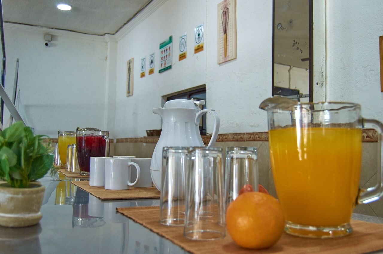 Uma mesa de café da manhã com diversas bebidas, um jarro, uma planta em vaso e um espelho refletindo o ambiente.
