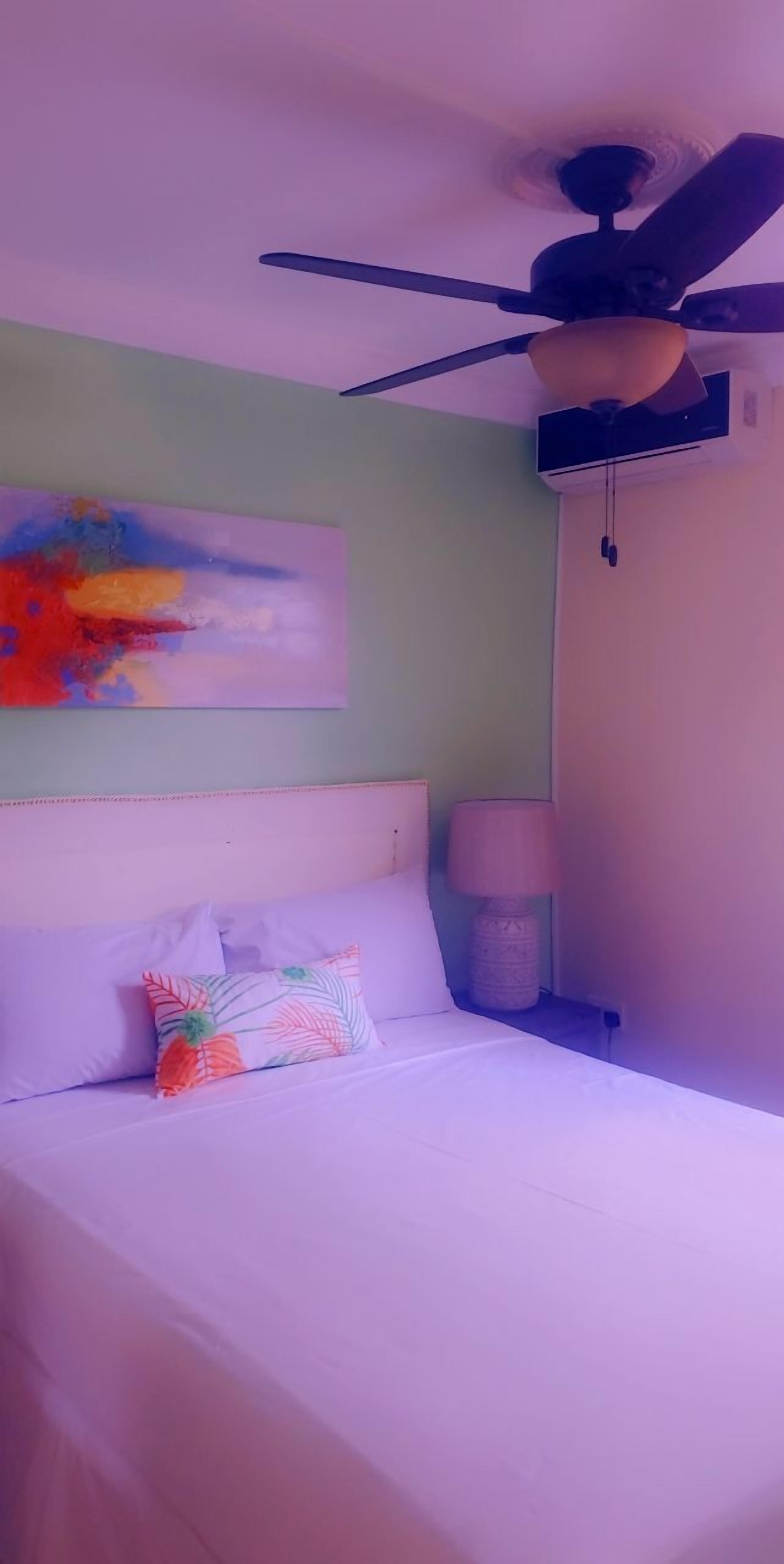Quarto com cama, ventilador de teto, ar condicionado fixado na parede e uma pintura abstrata colorida.