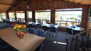 Um restaurante em estilo de cabana de madeira, com mesas e cadeiras, grandes janelas com vista para o lago, e um arranjo de flores na mesa.