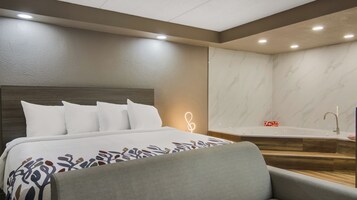 Quarto de hotel moderno com uma cama grande, um sofá cinzento, uma parede de mármore e um banheiro com banheira e um letreiro com a palavra 'LOVE'.