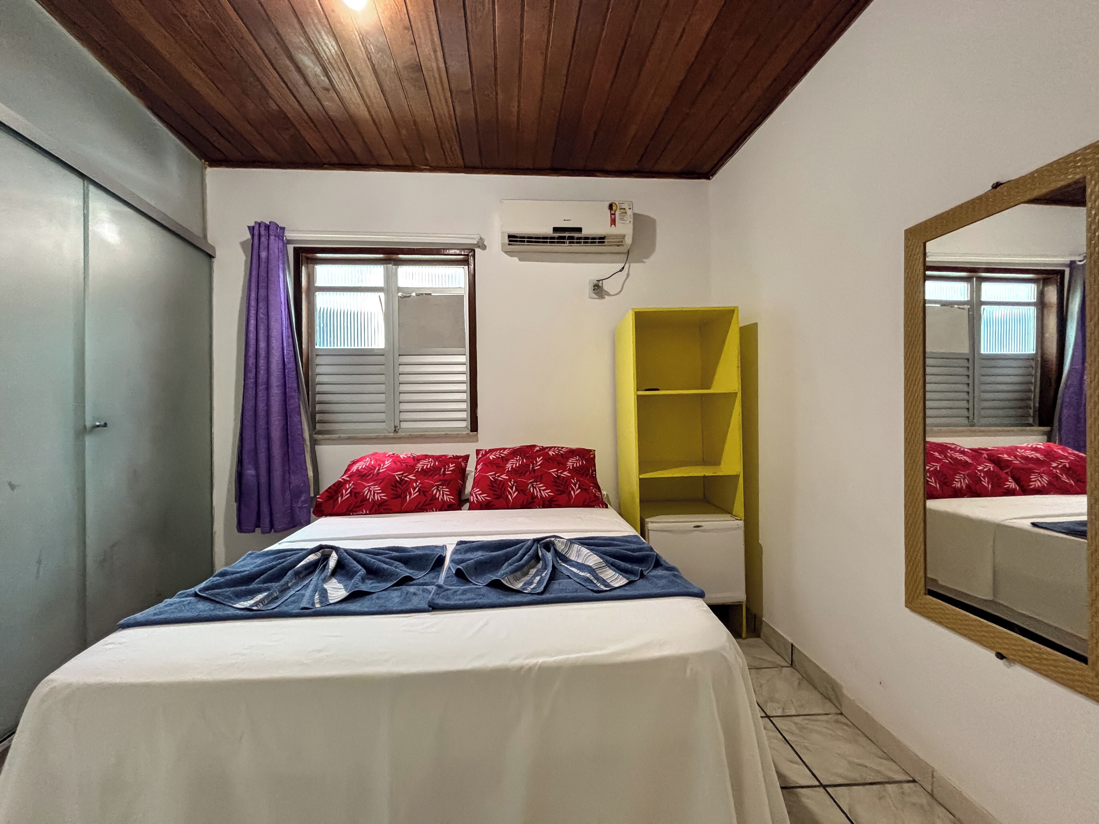 Quarto com cama, guarda-roupa, estante, ar condicionado e janela com cortinas.