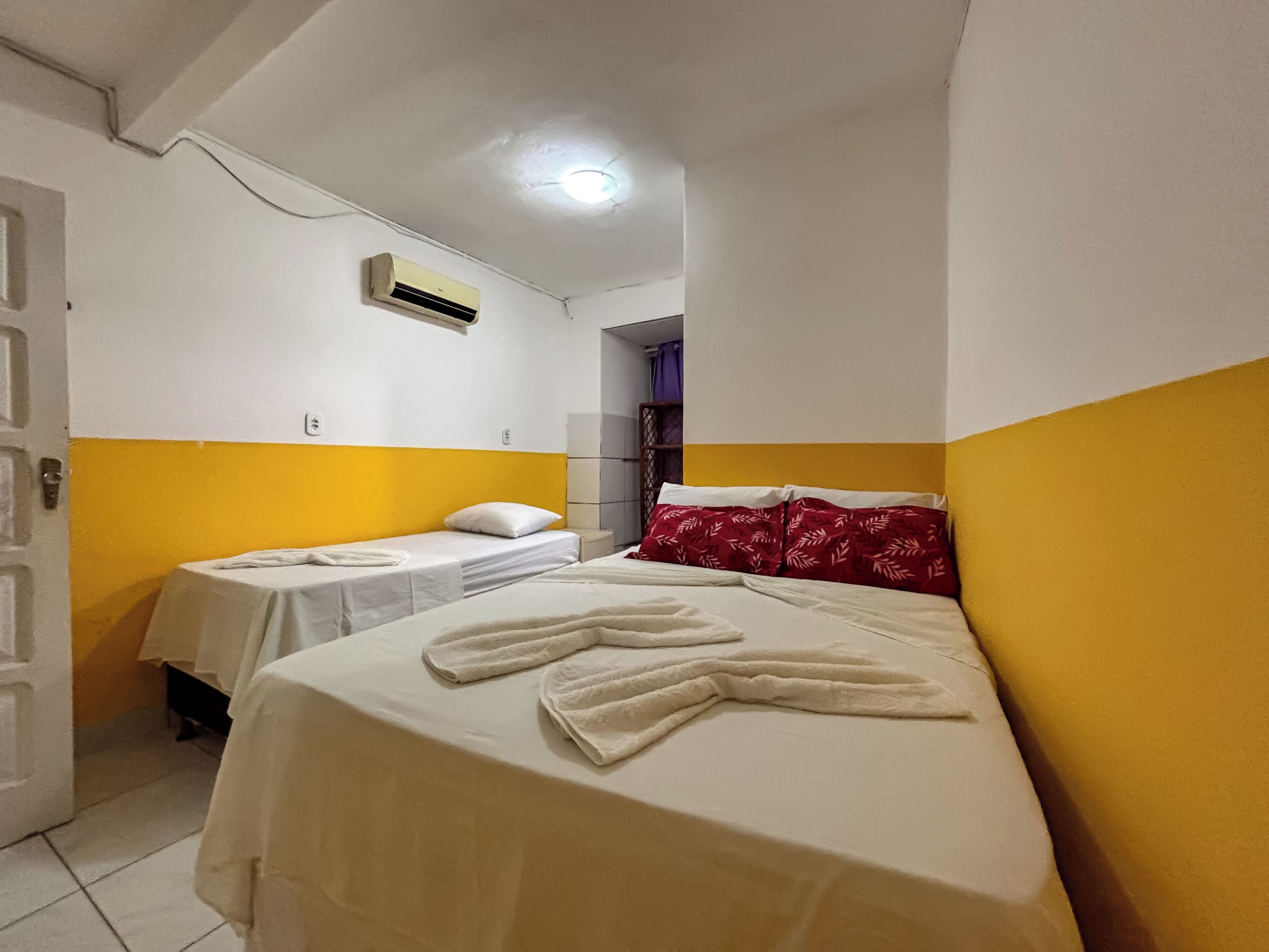 Quarto de hotel com duas camas, uma com colcha branca e outra com colcha estampada em vermelho e branco. Há um aparelho de ar condicionado branco na parede, e uma porta no lado esquerdo do quarto.