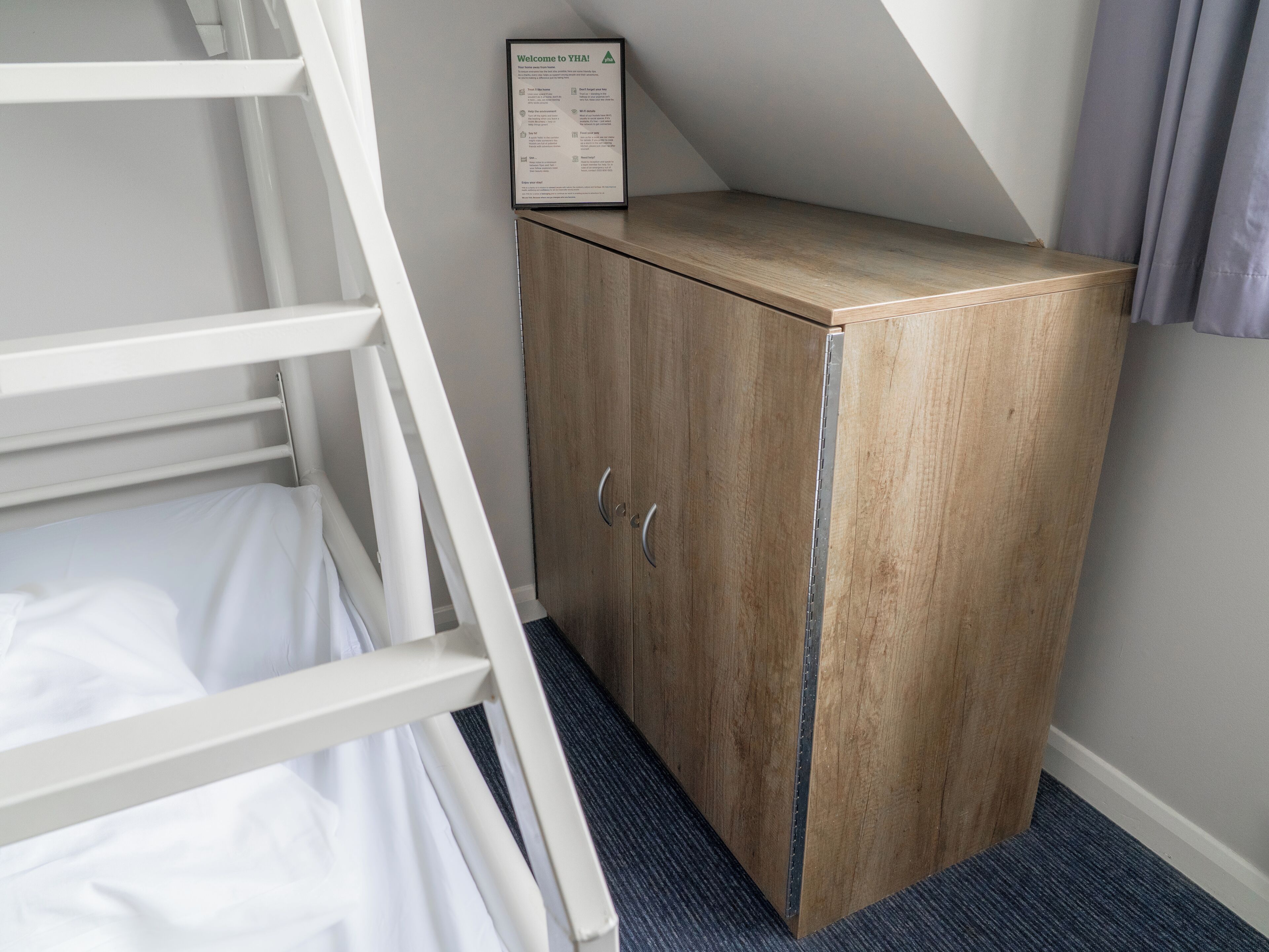 Uma cama beliche com um guarda-roupa de madeira ao lado, um letreiro de boas-vindas emoldurado e uma janela com cortinas.