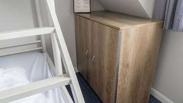 Uma cama beliche com um guarda-roupa de madeira ao lado, um letreiro de boas-vindas emoldurado e uma janela com cortinas.