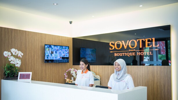 Uma receção de hotel com um ecrã digital, um arranjo floral e um aviso a indicar "SOVOTEL Boutique Hotel".