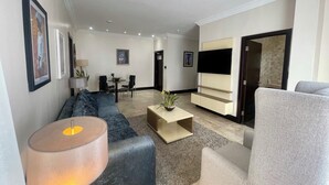 Uma sala de estar moderna com sofá, poltrona, mesa de centro e TV de tela plana na parede. Há também uma área de jantar com mesa e cadeiras, e uma porta que dá para outro cômodo.