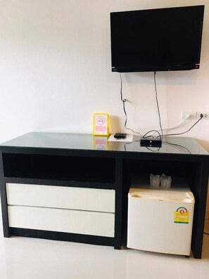 Quarto com escrivaninha preta, geladeira branca, TV fixada na parede e uma tomada.