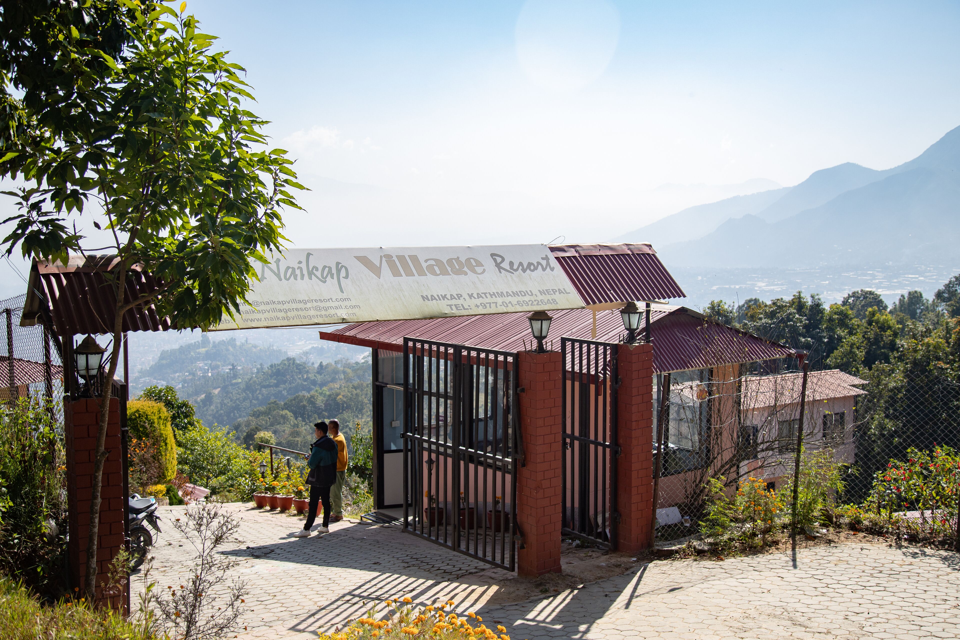 Entrada fechada com placa indicando Resort Naikap Village, com montanhas ao fundo.