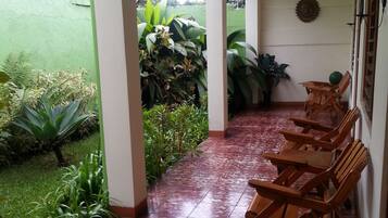 Uma área externa coberta com piso de azulejos, cadeiras de madeira e plantas em vasos.