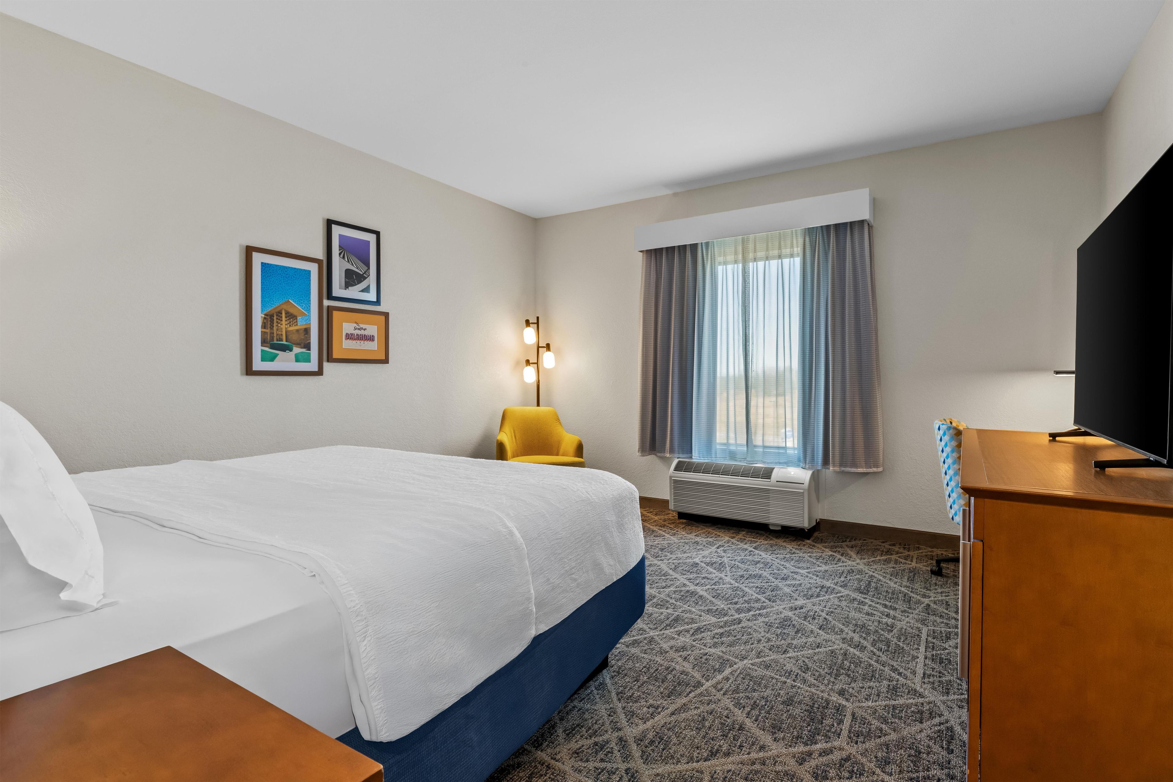 Quarto de hotel com cama, escrivaninha, cadeira, televisão, janela com cortinas e quadros nas paredes.