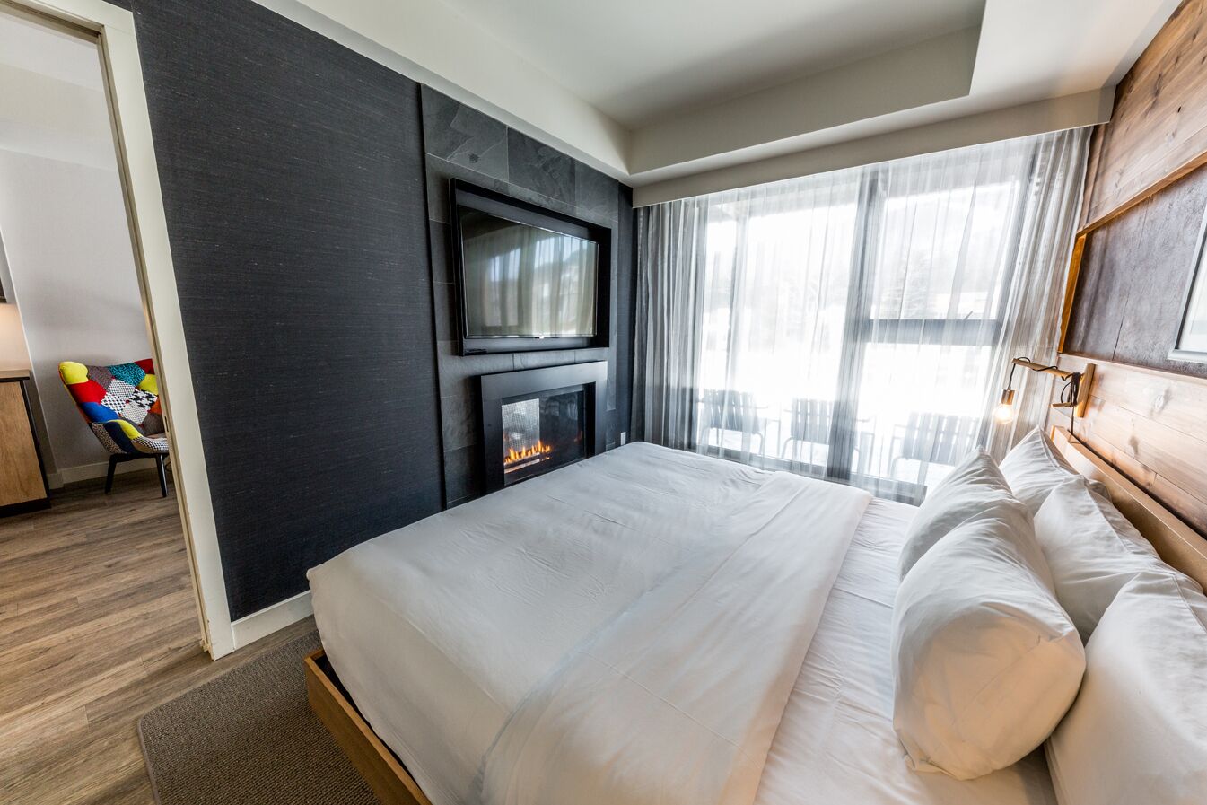 Quarto de hotel moderno com uma cama grande, lareira, televisão de tela plana e vista para o exterior através de cortinas finas.