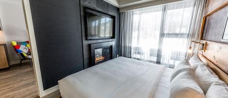 Quarto de hotel moderno com uma cama grande, lareira, televisão de tela plana e vista para o exterior através de cortinas finas.