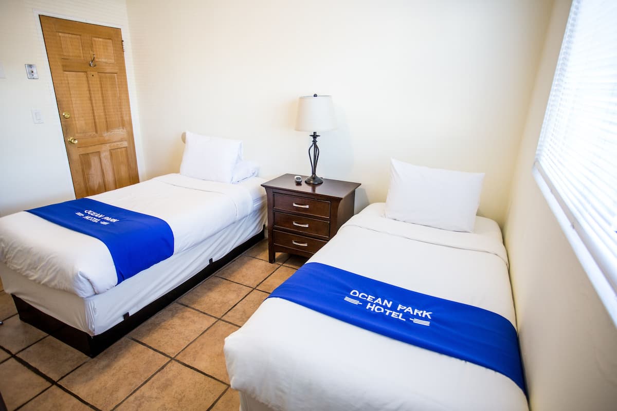 Quarto de hotel com duas camas, cada uma com um cobertor azul do Hotel Ocean Park, uma mesa de cabeceira com abajur e uma porta de madeira.