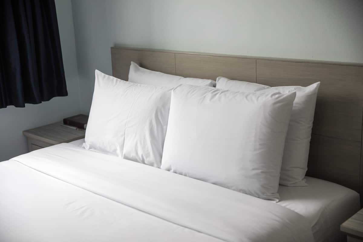 Uma cama bem arrumada com lençóis e almofadas brancas, uma cabeceira de madeira e uma mesa de cabeceira com abajur.