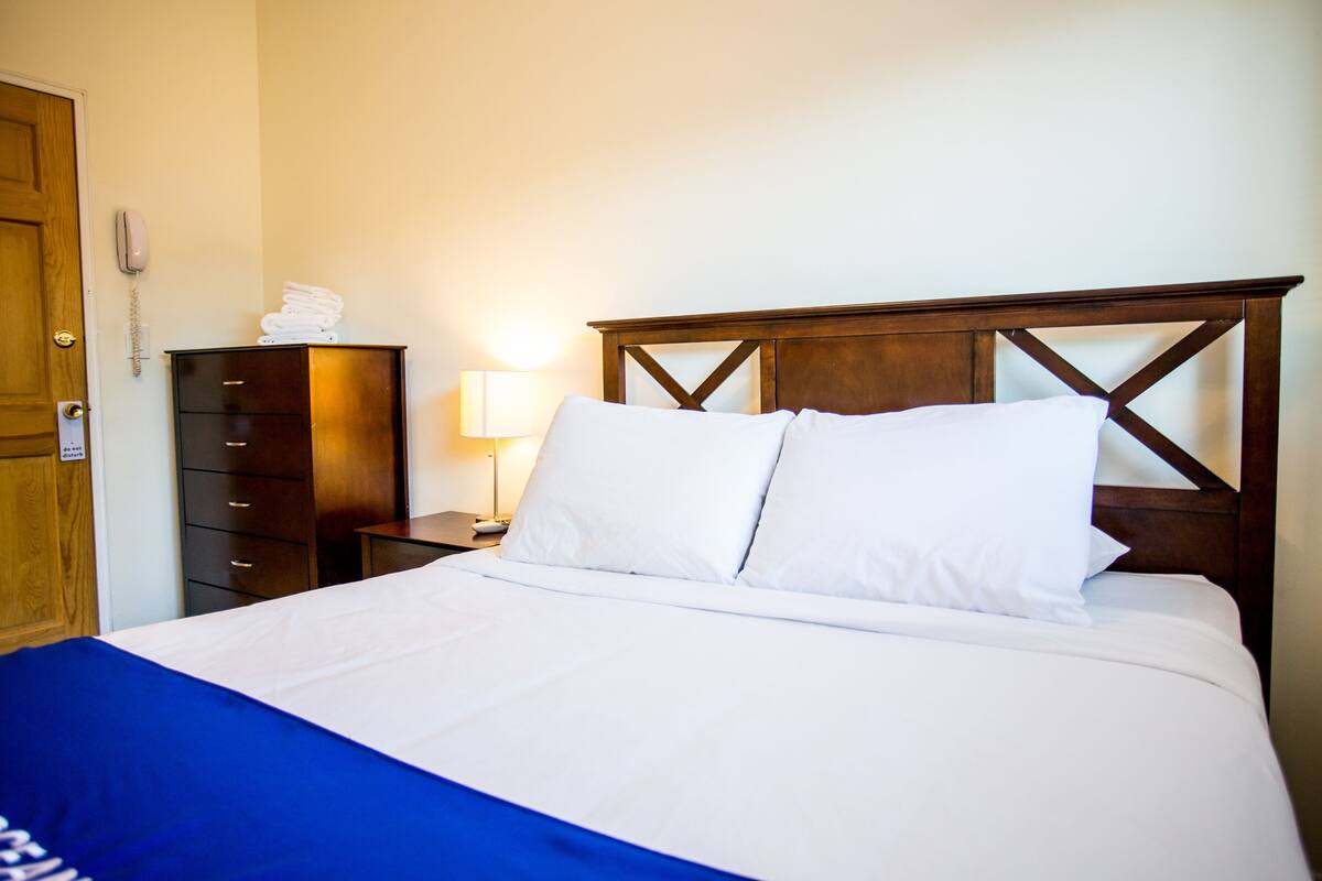 Uma cama bem arrumada com lençóis brancos e um toque de azul, um criado-mudo de madeira com uma luminária, e um guarda-roupa de madeira com um telefone em cima.