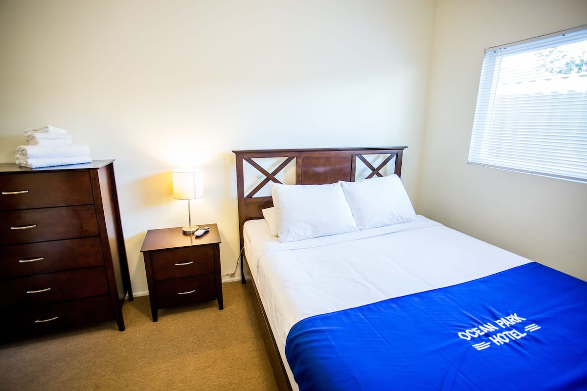 Quarto de hotel com cama, mesa de cabeceira com luminária, cômoda e janela com persianas.