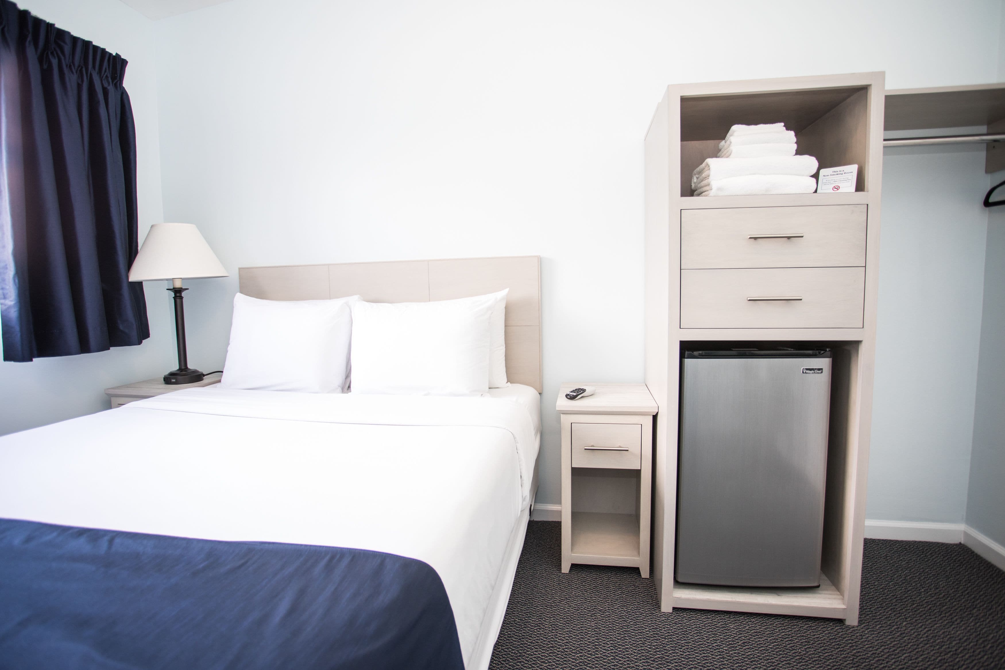 Quarto de hotel com cama, mesa de cabeceira, luminária, armário e geladeira.