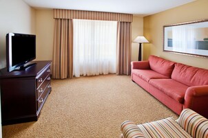 Quarto de hotel com televisão em uma cômoda, um sofá vermelho, uma cadeira listrada, um abajur e uma janela grande com cortinas.