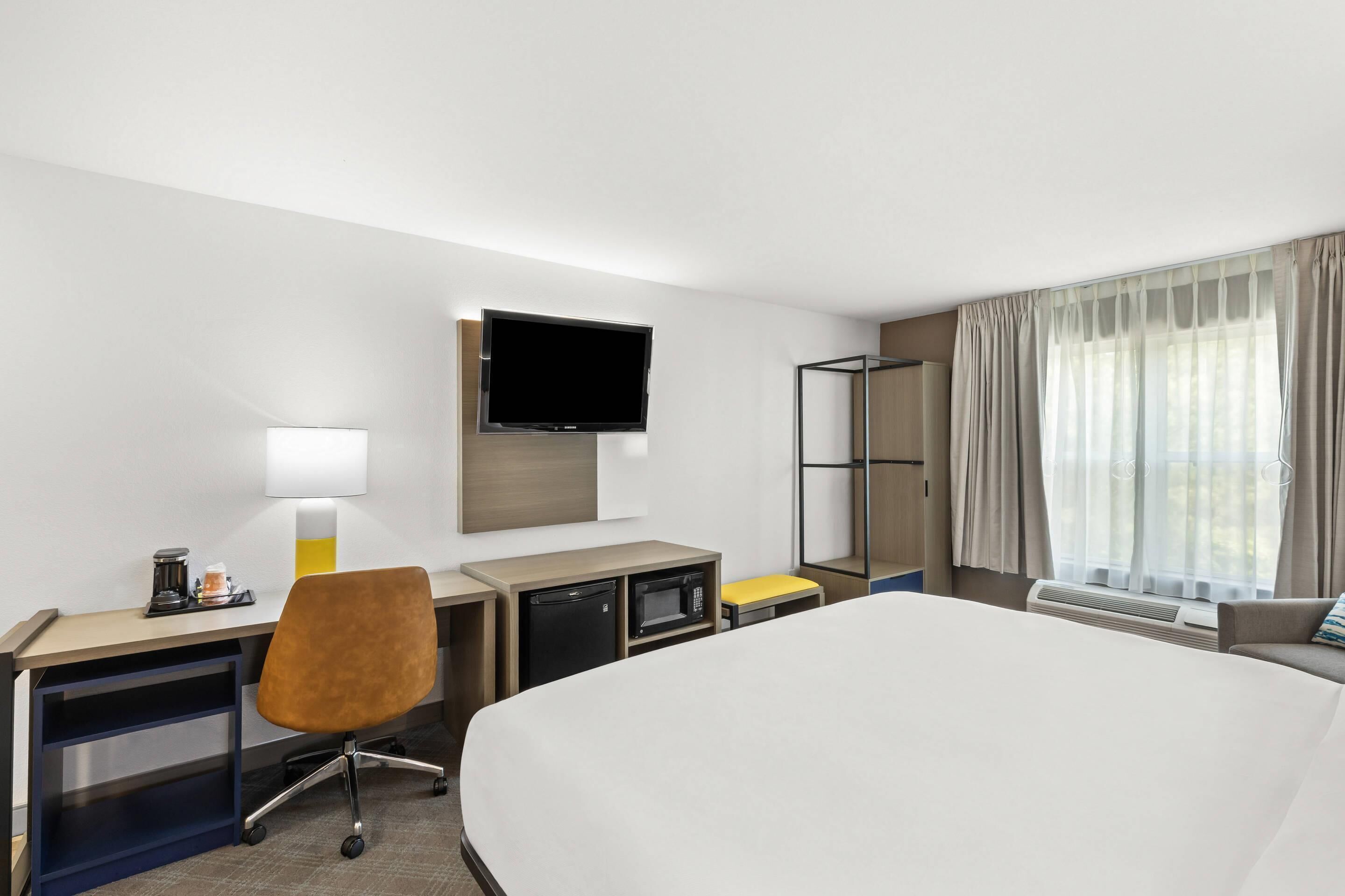 Quarto de hotel com cama, escrivaninha, cadeira, televisão e janela com cortinas.