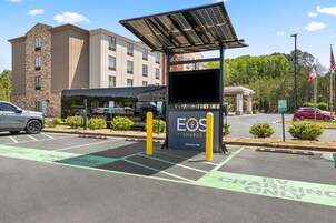 Uma área de estacionamento com estações de carregamento para veículos elétricos, um edifício de hotel e um mastro com bandeiras.