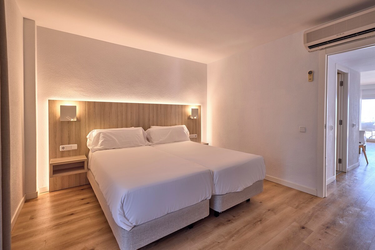 Cama de casal com cabeceira de madeira e abajures, piso de madeira e uma porta que dá para outro quarto.