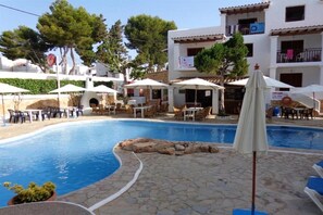 Uma área de piscina com espreguiçadeiras e guarda-sóis, cercada por um terraço de pedra e um edifício com varandas.