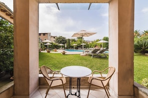 Um pátio com uma mesa redonda e cadeiras de vime, com vista para uma área de piscina com espreguiçadeiras e guarda-sóis.