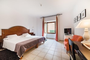 Quarto de hotel com cama de madeira, uma escrivaninha com luminária, uma cadeira e uma televisão.