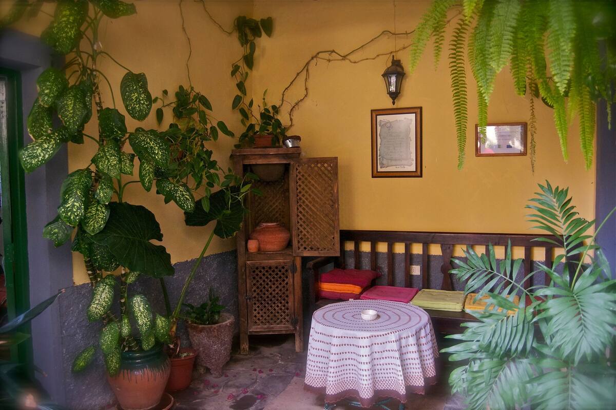 Um recanto acolhedor com um banco de madeira, uma mesinha redonda com toalha de renda, plantas em vasos e um candeeiro fixado na parede.