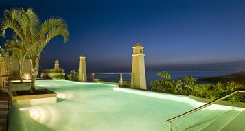 Uma piscina com vista para o oceano à noite, rodeada por palmeiras e um edifício com uma torre.