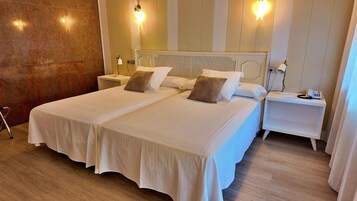 Uma cama de casal com roupa de cama branca e duas almofadas, uma mesa de cabeceira com abajur e um aplique de parede.
